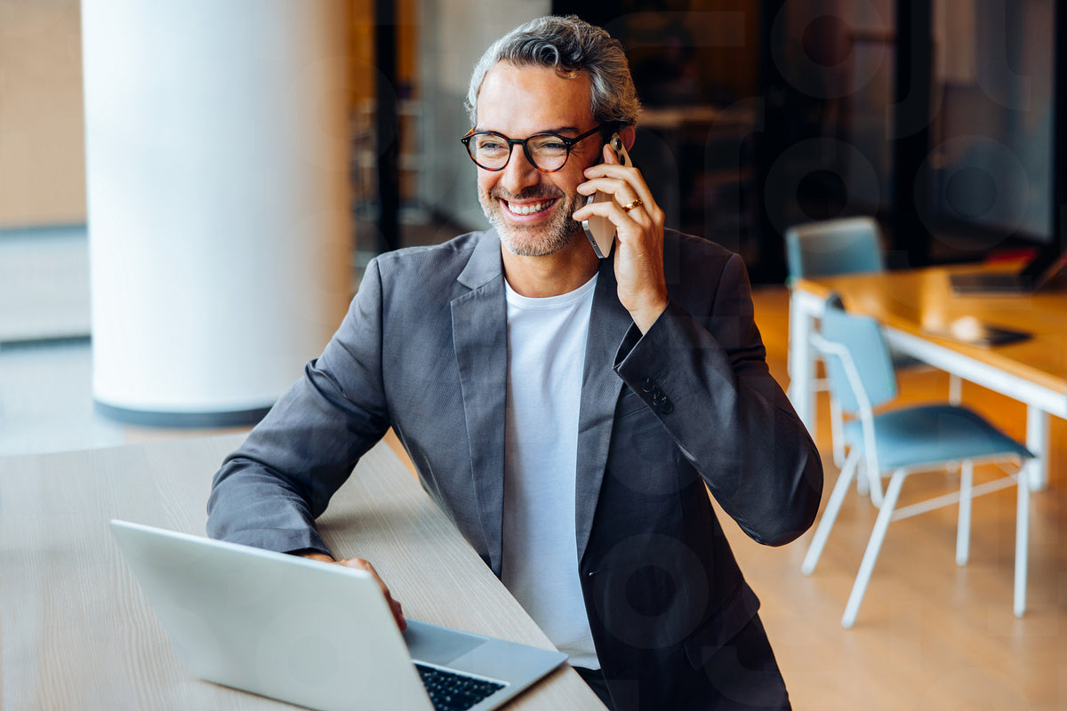 Professional man using a phone while working on a laptop at an office ...