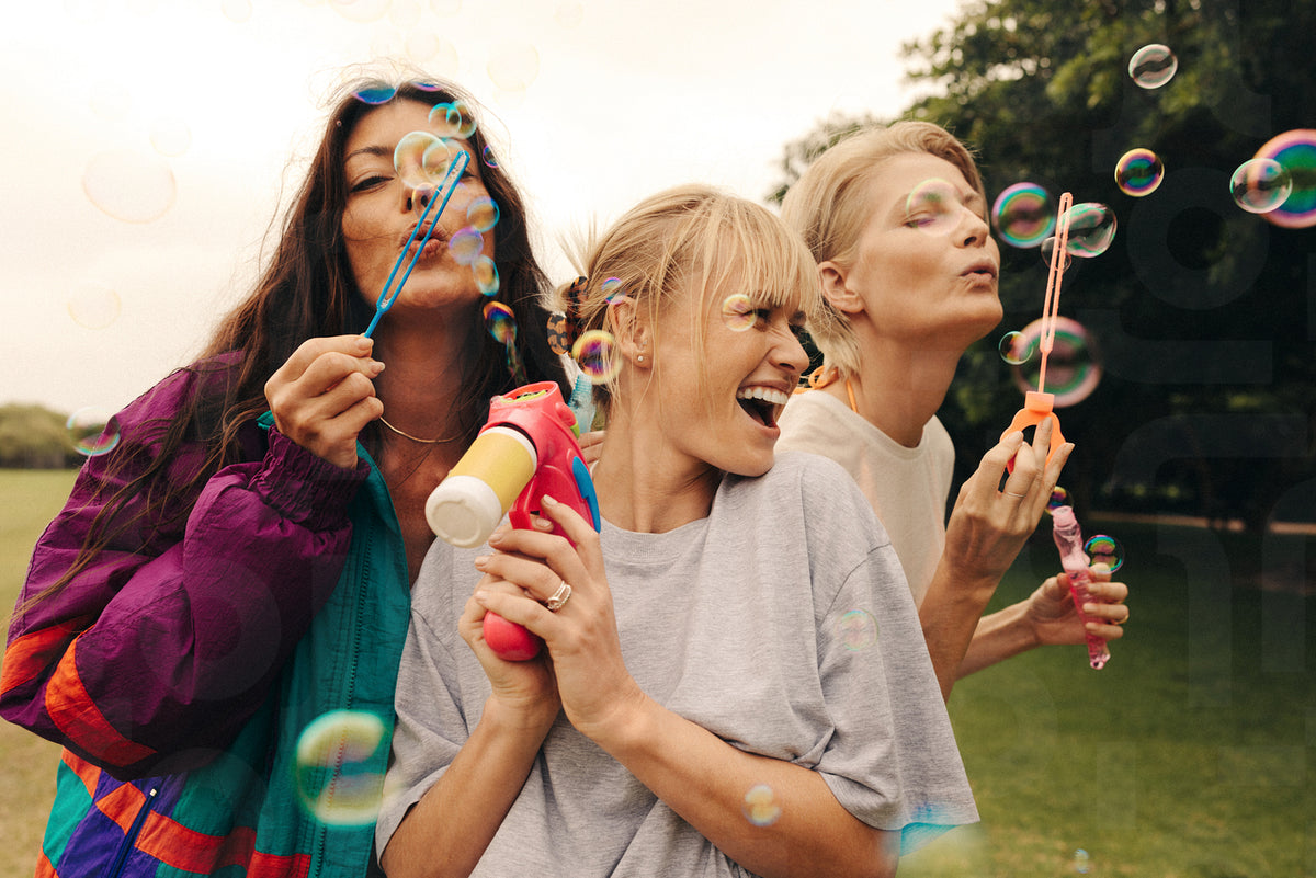 Three carefree female friends have fun blowing bubbles in a park ...
