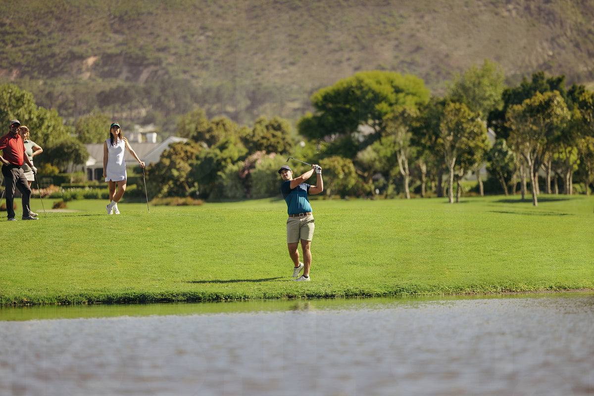 Male golfer swinging golf club near water hazard with team watching ...